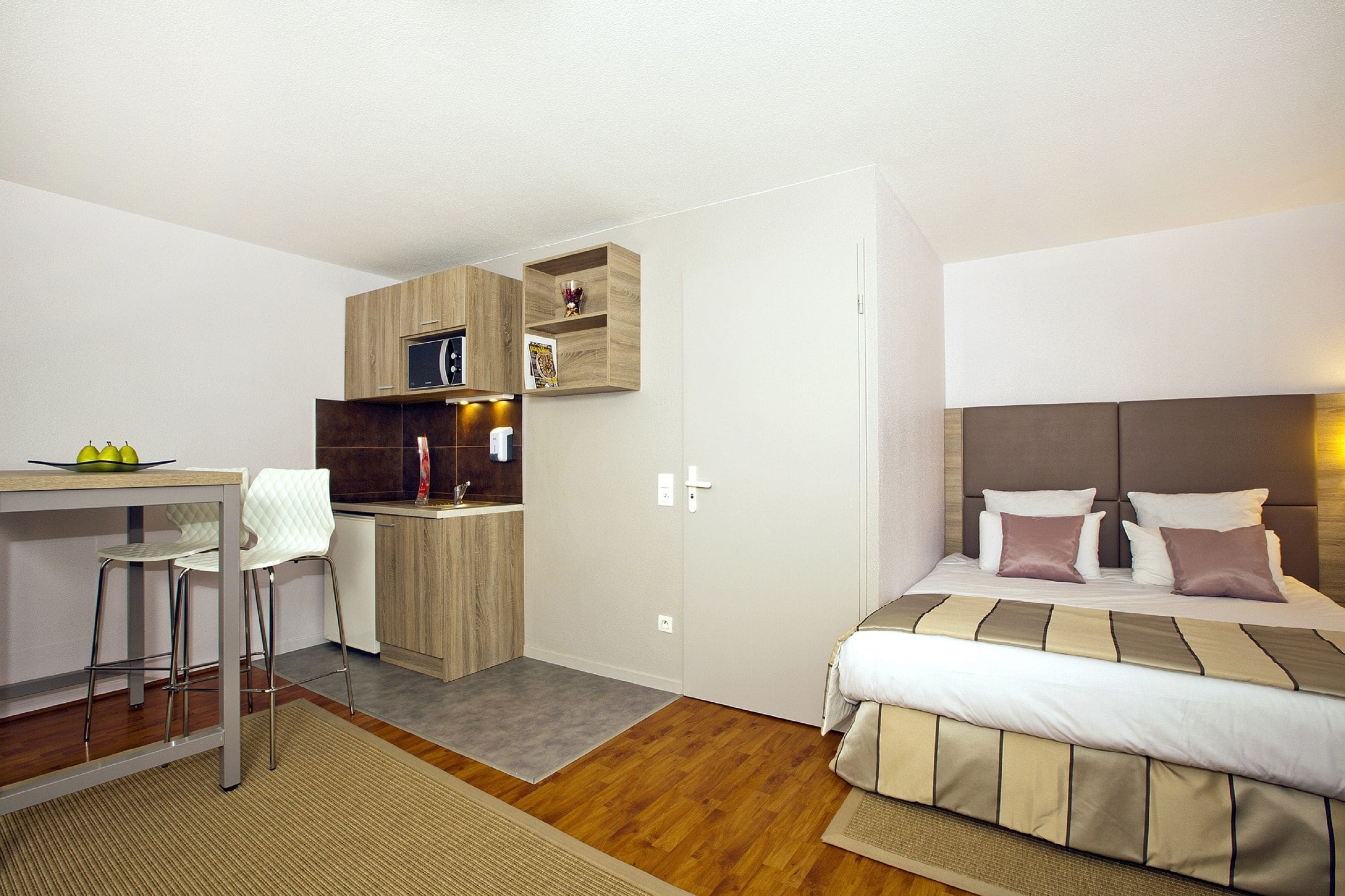 Quarto de hotel compacto com cozinha americana, cama com cabeceira e pequena área de jantar.