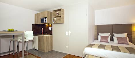 Quarto de hotel compacto com cozinha americana, cama com cabeceira e pequena área de jantar.