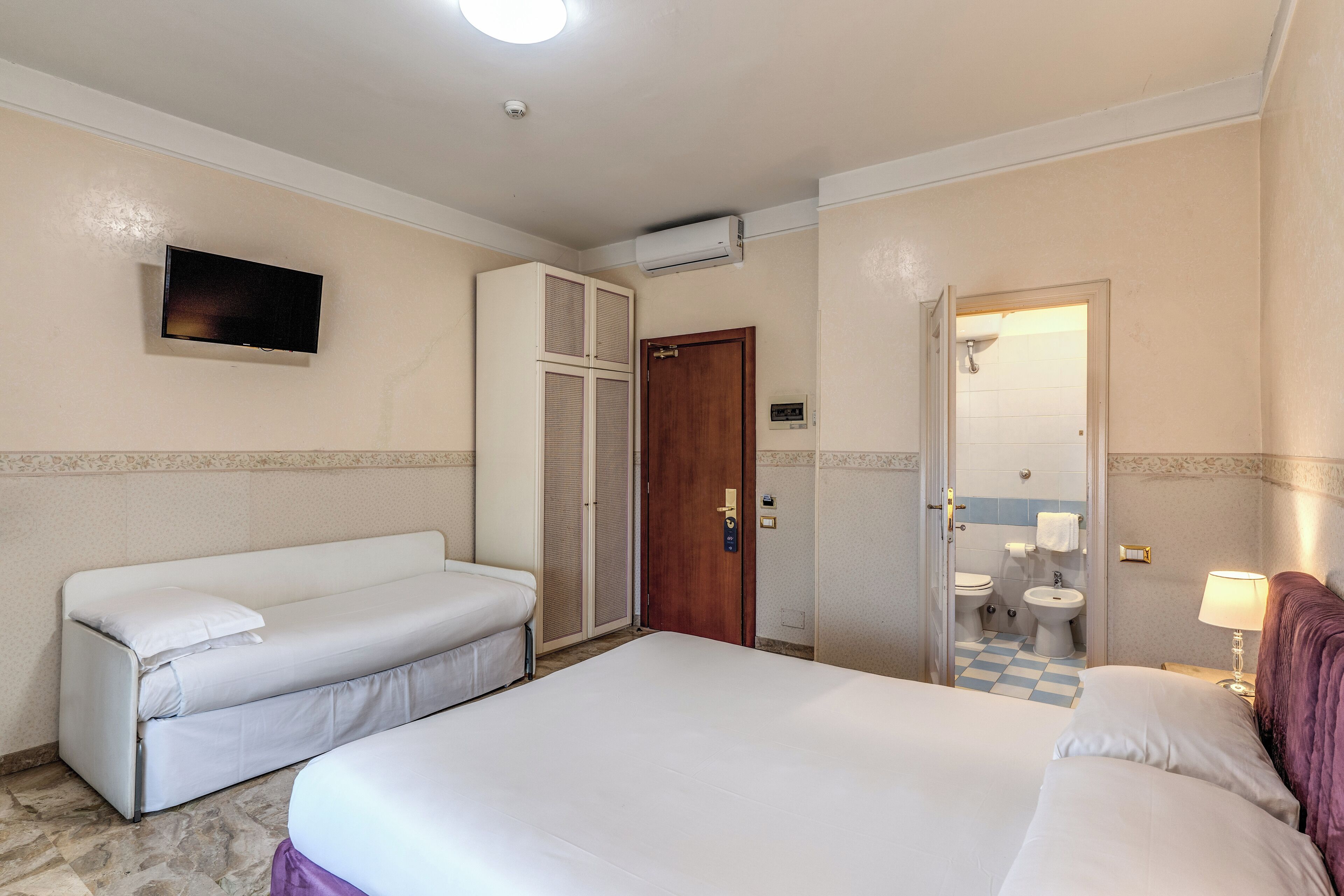 Quarto de hotel com uma cama, um sofá, uma televisão, ar condicionado e um banheiro visível por uma porta aberta.