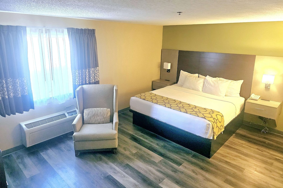 Quarto de hotel com cama, uma cadeira, mesa de cabeceira, abajur e janela com cortinas.