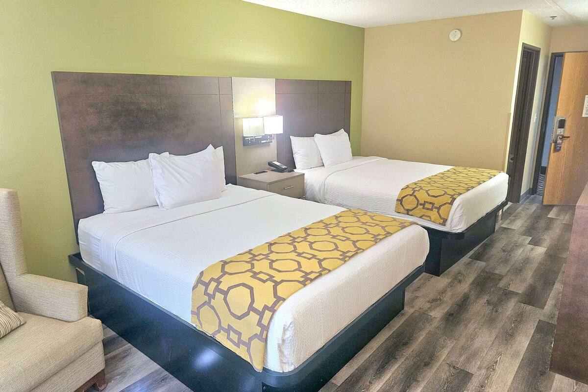 Quarto de hotel com duas camas, uma mesinha de cabeceira, uma cadeira e uma porta.