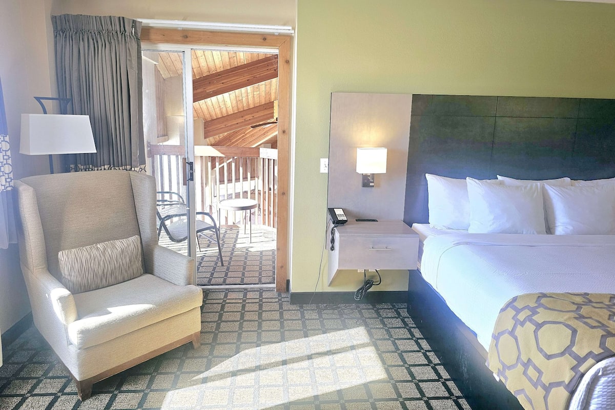 Quarto de hotel com cama, uma cadeira, escrivaninha com luminária e vista para área externa.