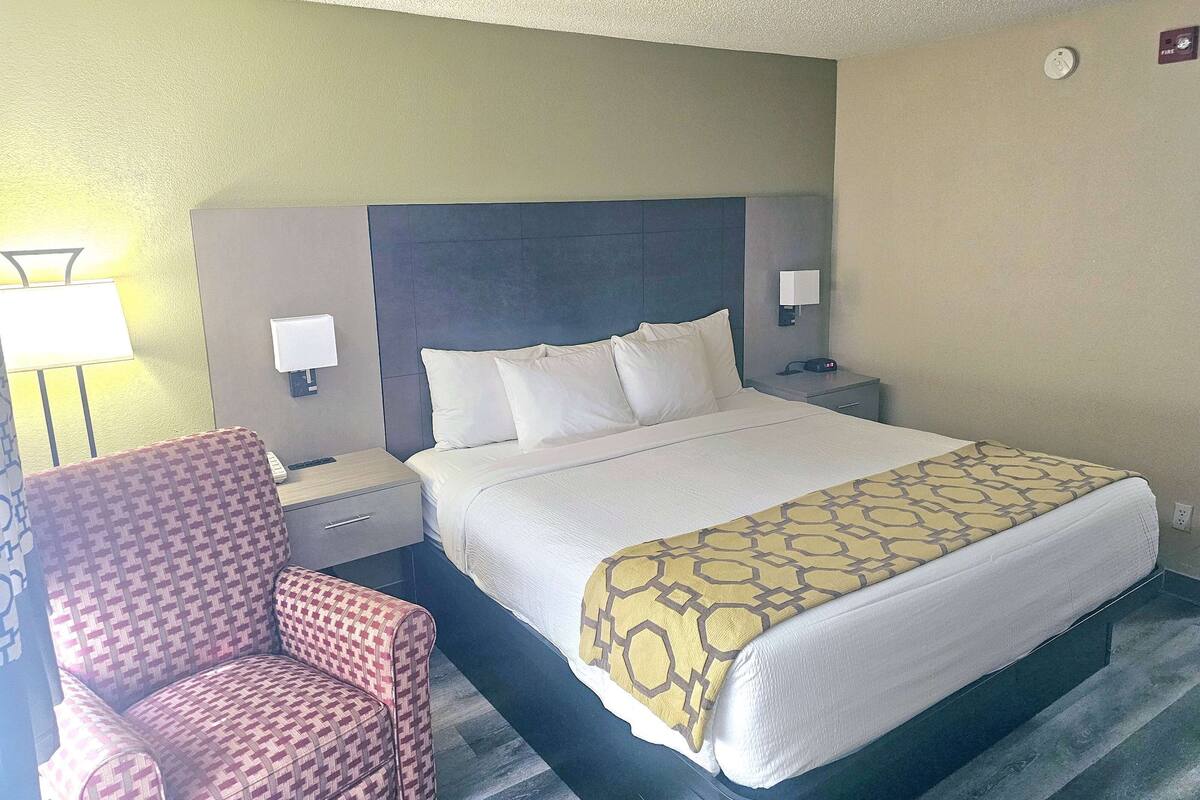 Quarto de hotel com uma cama grande, uma poltrona estampada, mesas de cabeceira com abajures e uma cabeceira.