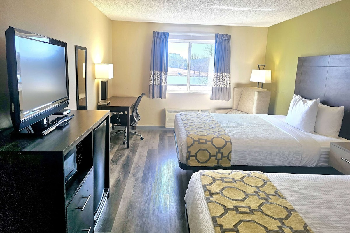 Quarto de hotel com televisão, escrivaninha, duas camas e janela com cortinas.