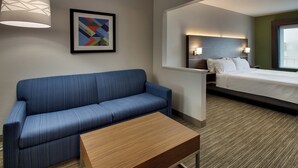 Quarto de hotel com sofá azul, mesa de centro de madeira, cama com roupa de cama branca, cabeceira com luzes fixadas na parede e uma pintura abstrata emoldurada.