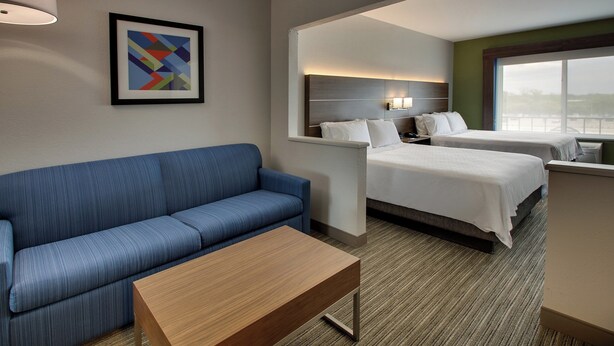 Quarto de hotel com sofá azul, mesa de madeira, duas camas, obra de arte abstrata emoldurada e uma janela grande.