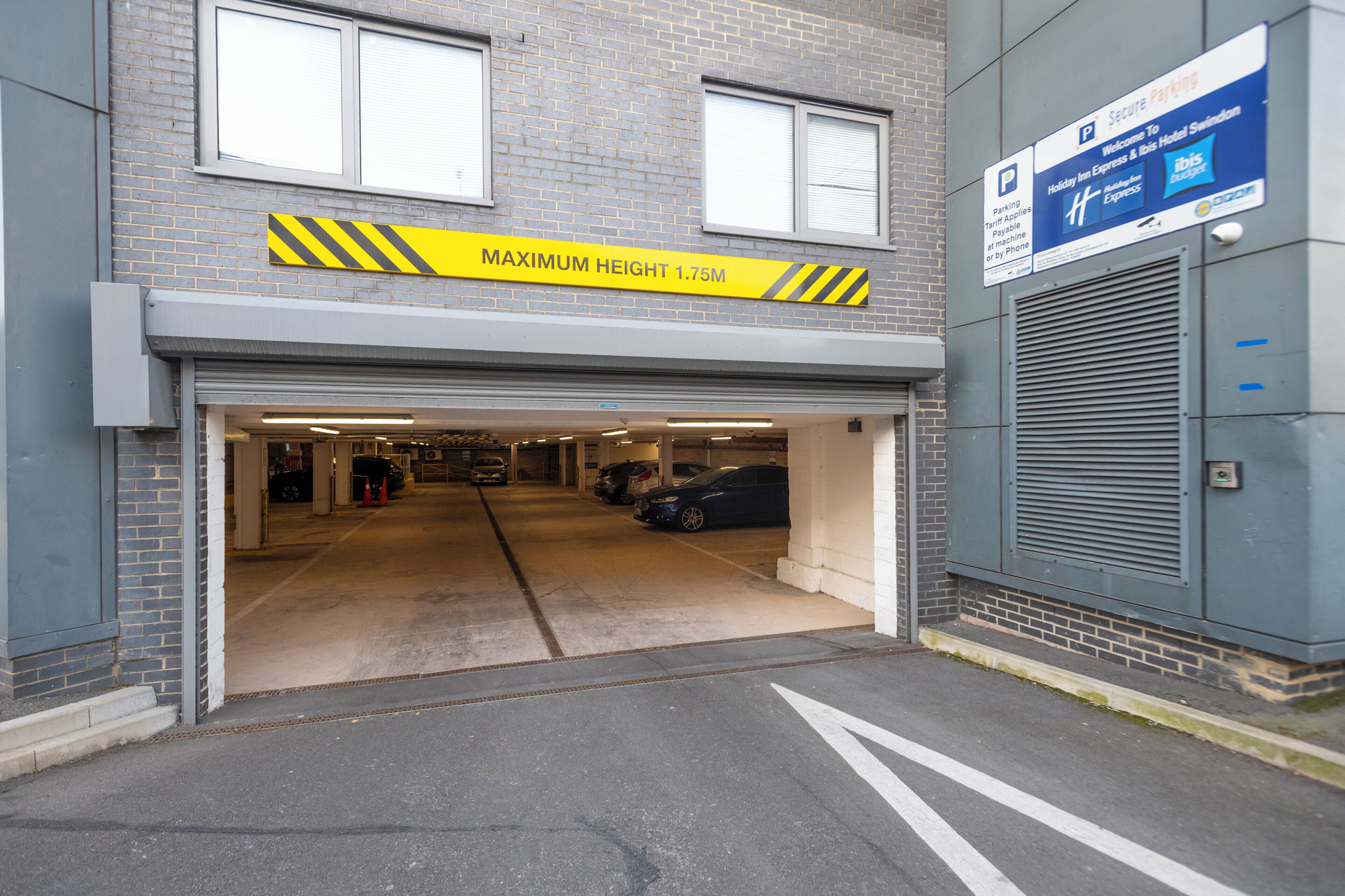 Entrada de um estacionamento com placa de altura máxima, sinalização de Holiday Inn Express e Ibis Hotel, e placa indicando estacionamento seguro.