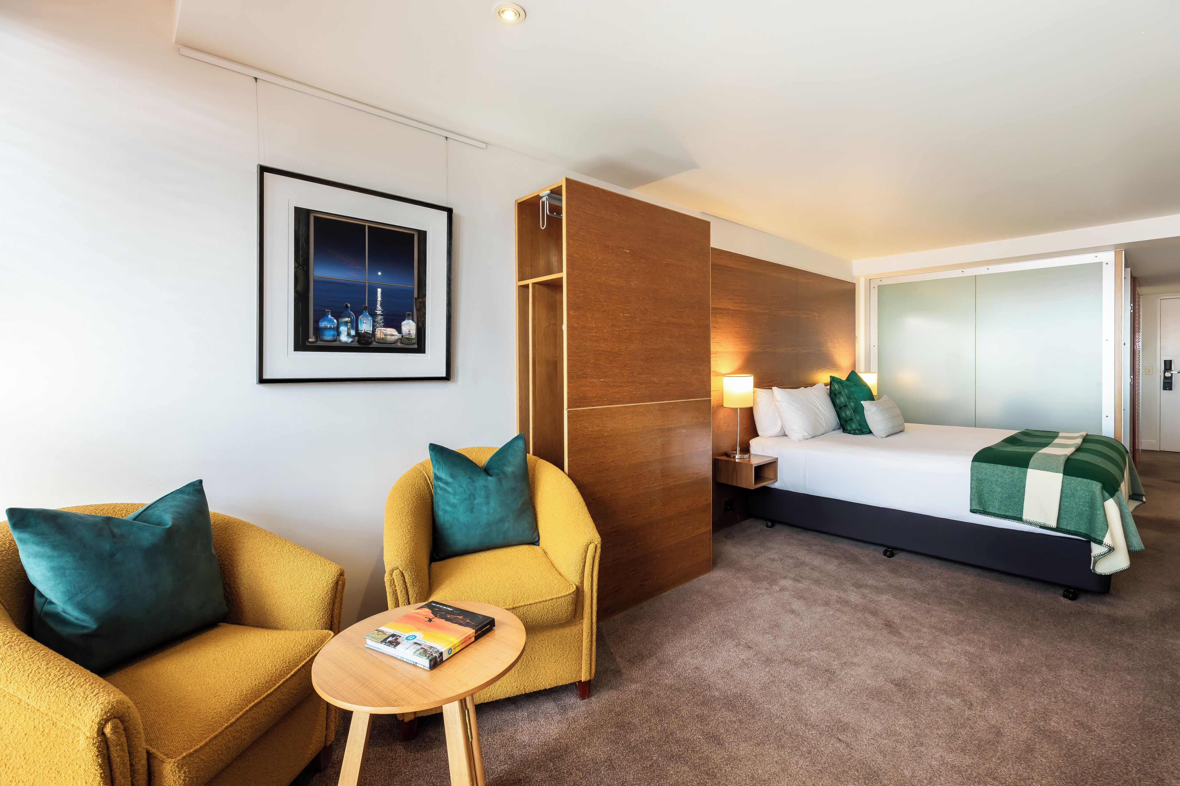 Quarto de hotel com uma cama, duas poltronas, uma mesinha, criado-mudo, luminária e um quadro na parede.