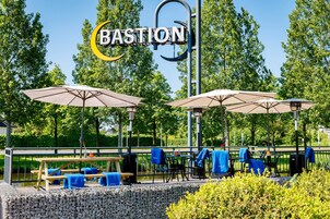 Uma área externa com mesas, guarda-sóis, uma placa com a inscrição "BASTION" e vista para árvores e um corpo d'água.