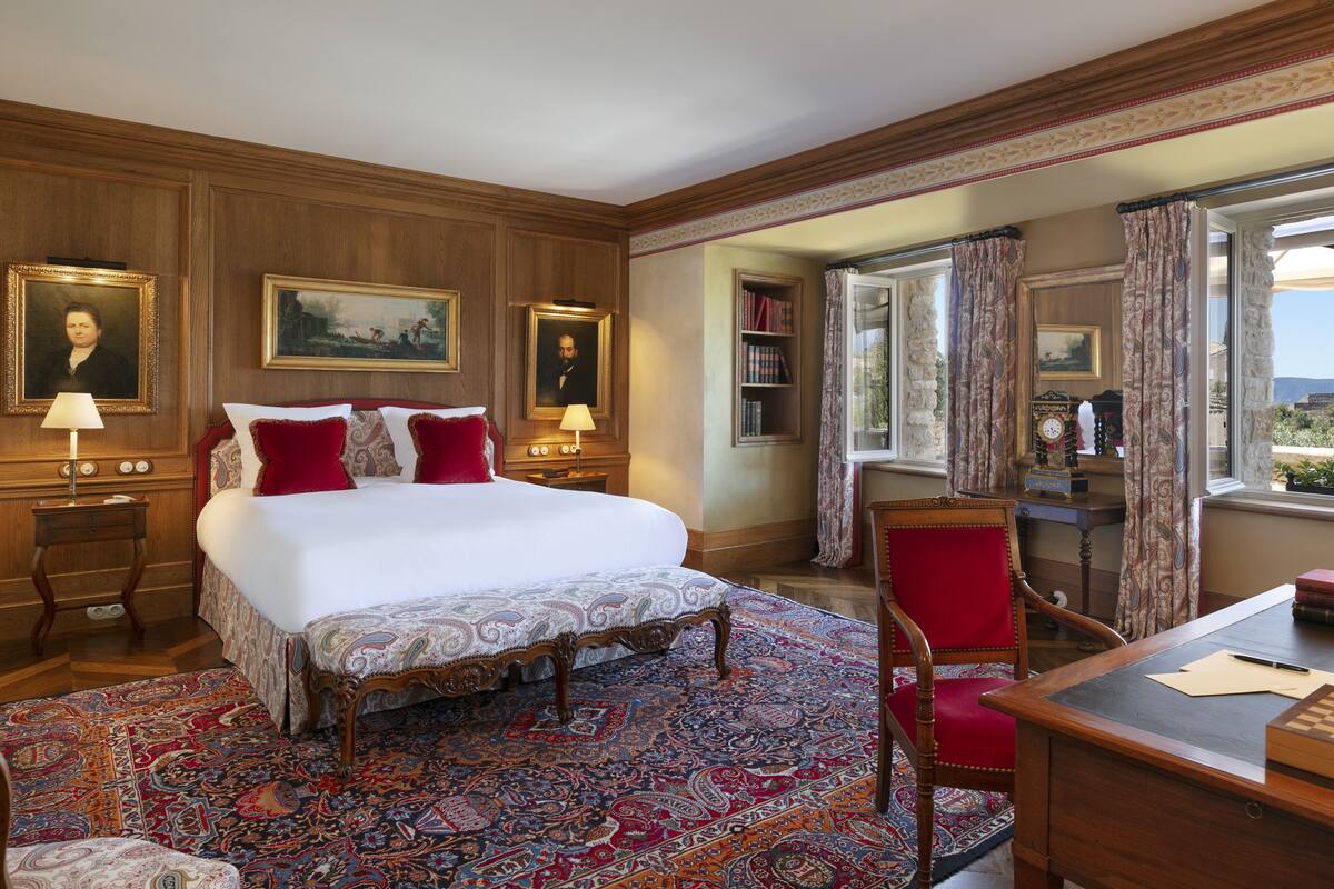 Quarto de hotel com uma cama grande, duas mesas de cabeceira com luminárias, uma cadeira vermelha, uma escrivaninha com um tabuleiro de xadrez e vista para o exterior através de uma janela.
