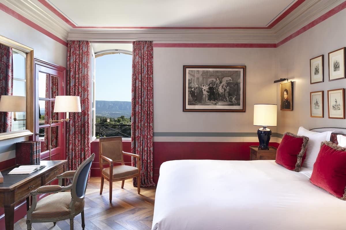 Quarto de hotel com uma cama grande, uma escrivaninha com cadeira, uma janela com cortinas e um quadro na parede.