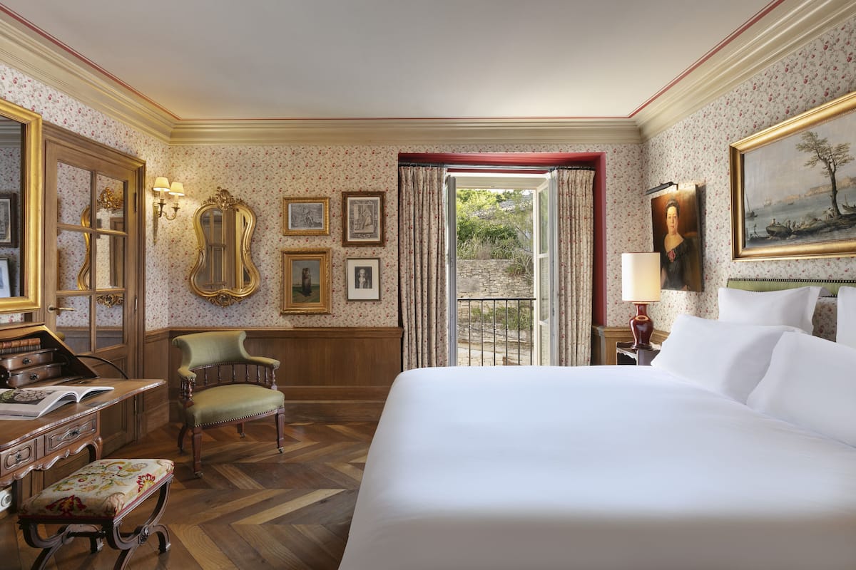 Quarto de hotel com uma cama grande, uma escrivaninha de madeira com cadeira, uma mesinha com um banco e vista para o exterior através de uma porta de vidro.