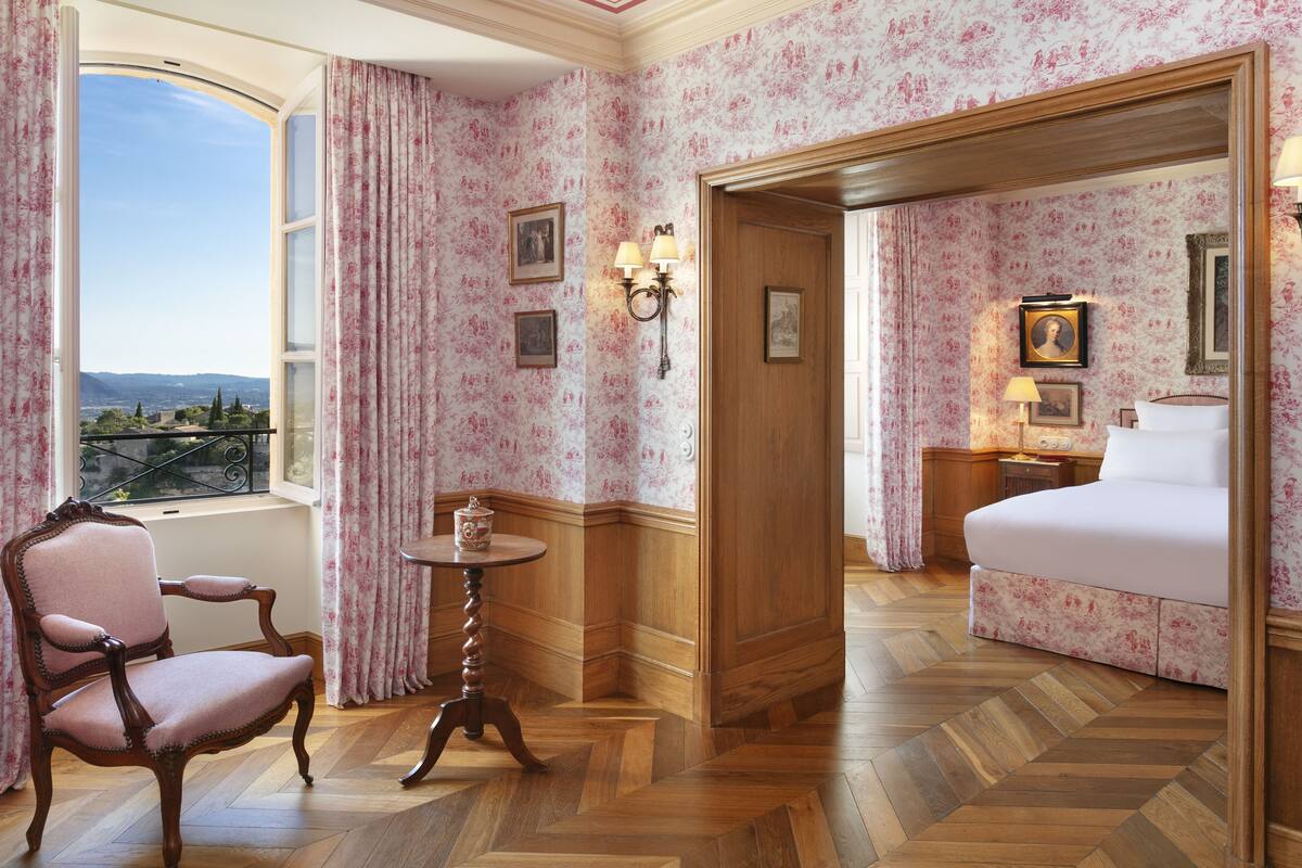 Quarto de hotel com piso de madeira, uma cama, uma cadeira, uma mesa com abajur e vista para o exterior.