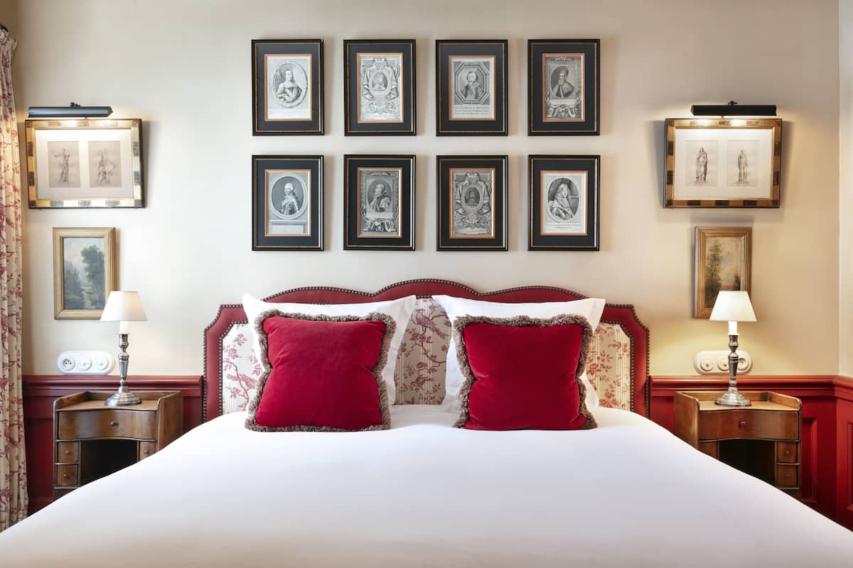 Quarto de hotel com uma cama grande, roupa de cama vermelha e branca, duas mesas de cabeceira com abajures e uma parede decorada com retratos emoldurados.