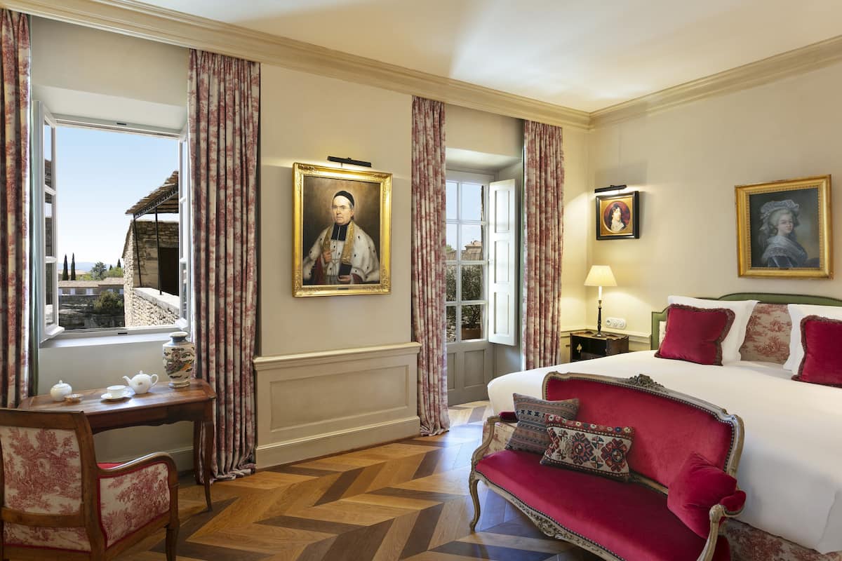 Quarto de hotel com uma cama grande, uma escrivaninha com cadeira, uma janela com cortinas e um quadro na parede.