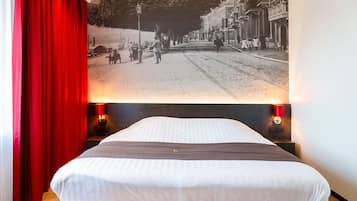 Quarto de hotel com uma cama grande, cortinas vermelhas, cabeceira com luzes fixadas na parede e um cartaz de uma cena de rua antiga.