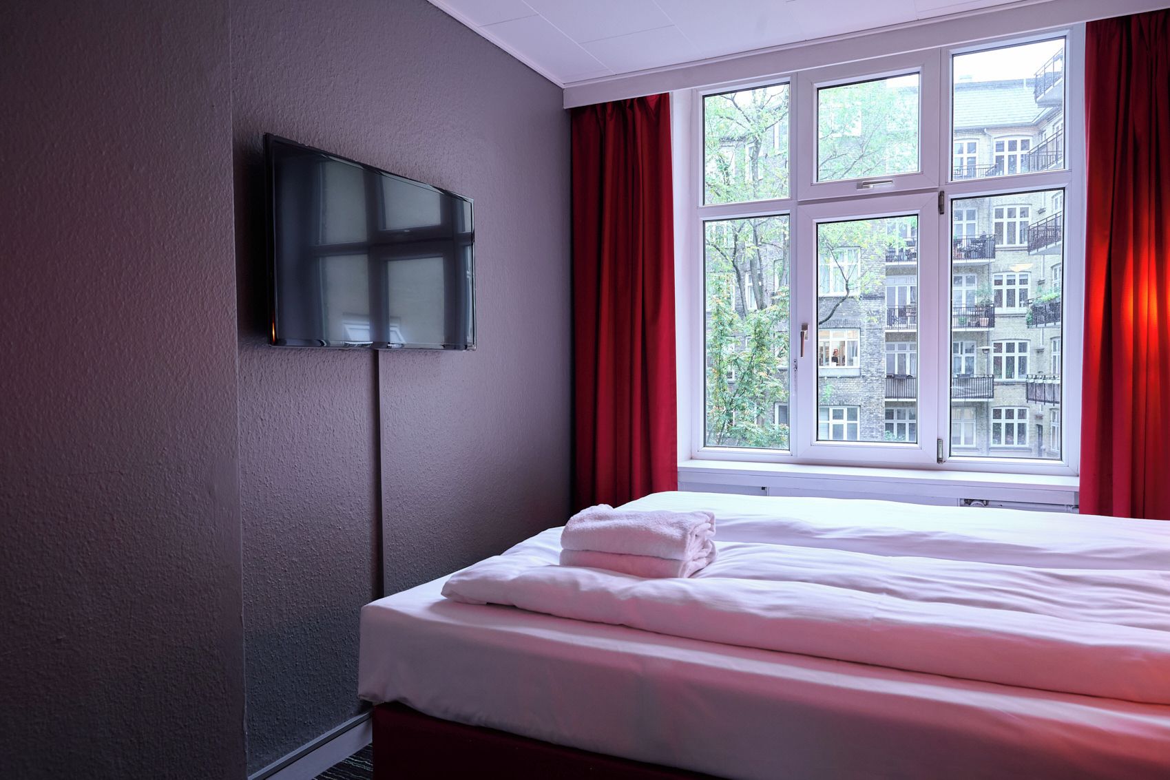 Quarto de hotel com cama, televisão de tela plana na parede e janela com cortinas vermelhas.