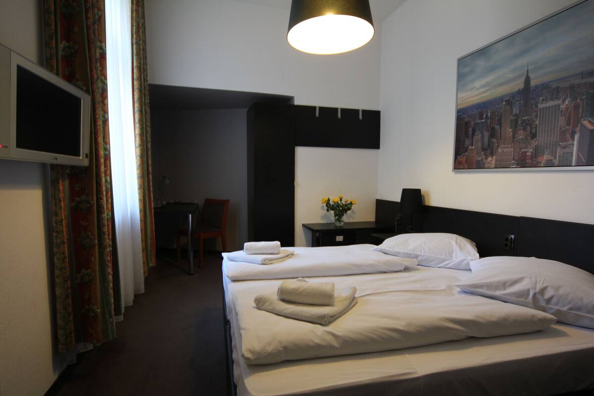 Quarto de hotel com cama grande, escrivaninha, cadeira, pintura de paisagem urbana e televisão na parede.