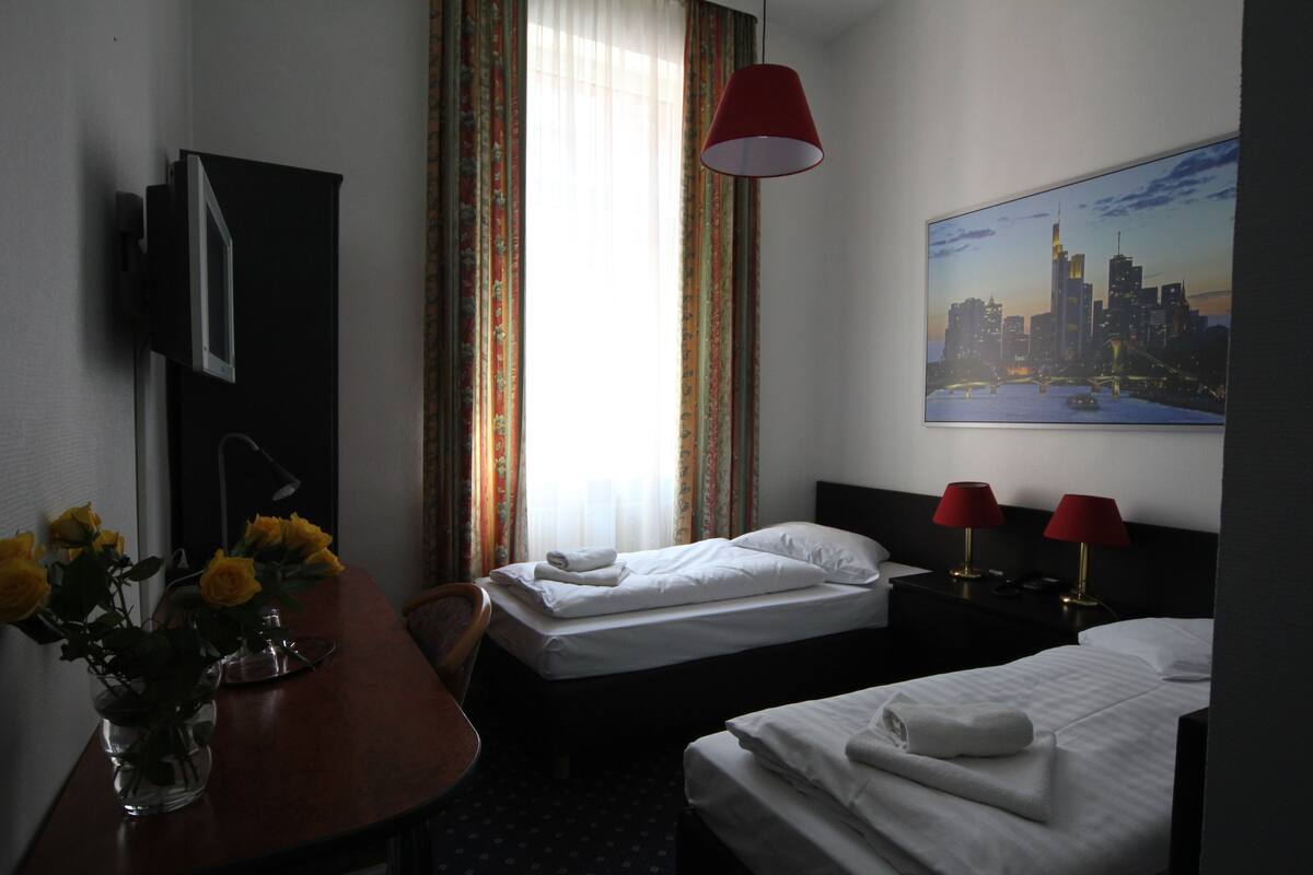 Quarto de hotel com duas camas, uma escrivaninha com cadeira, um vaso com flores, um quadro de uma paisagem urbana e um abajur vermelho.
