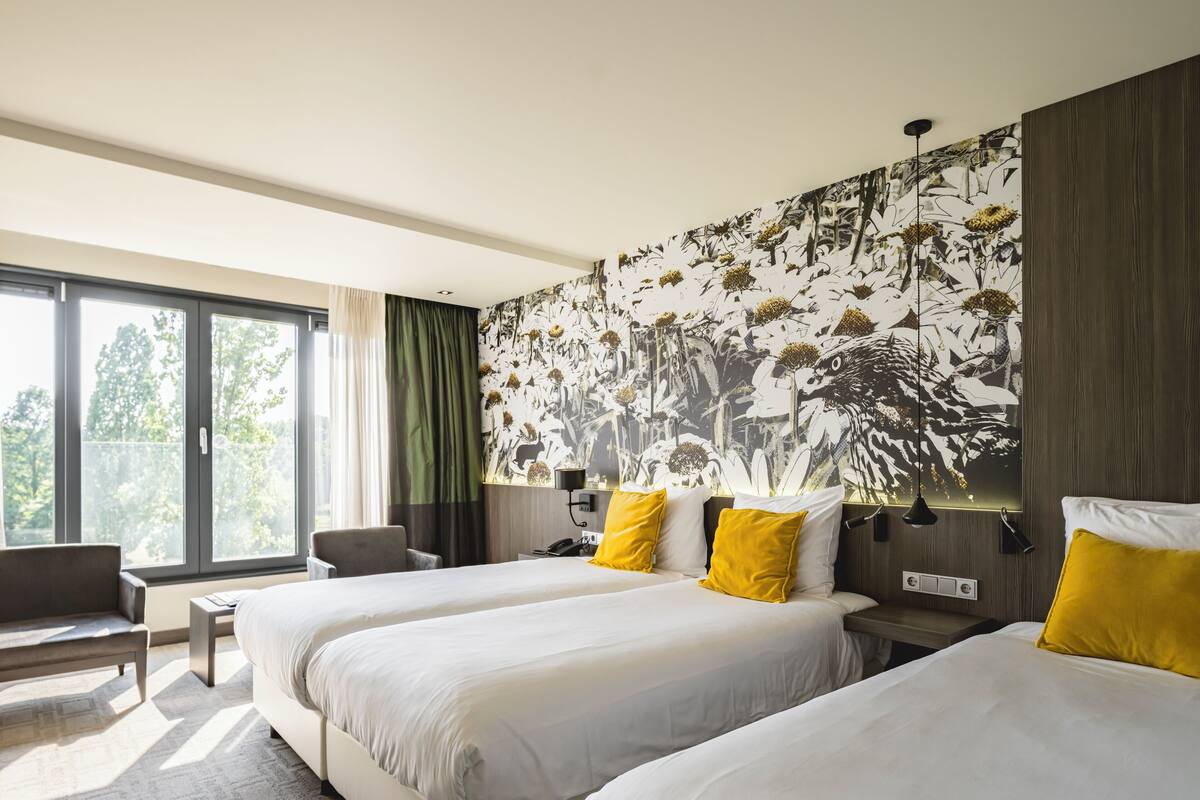 Quarto de hotel com duas camas, uma escrivaninha, uma cadeira e uma janela grande com cortinas.
