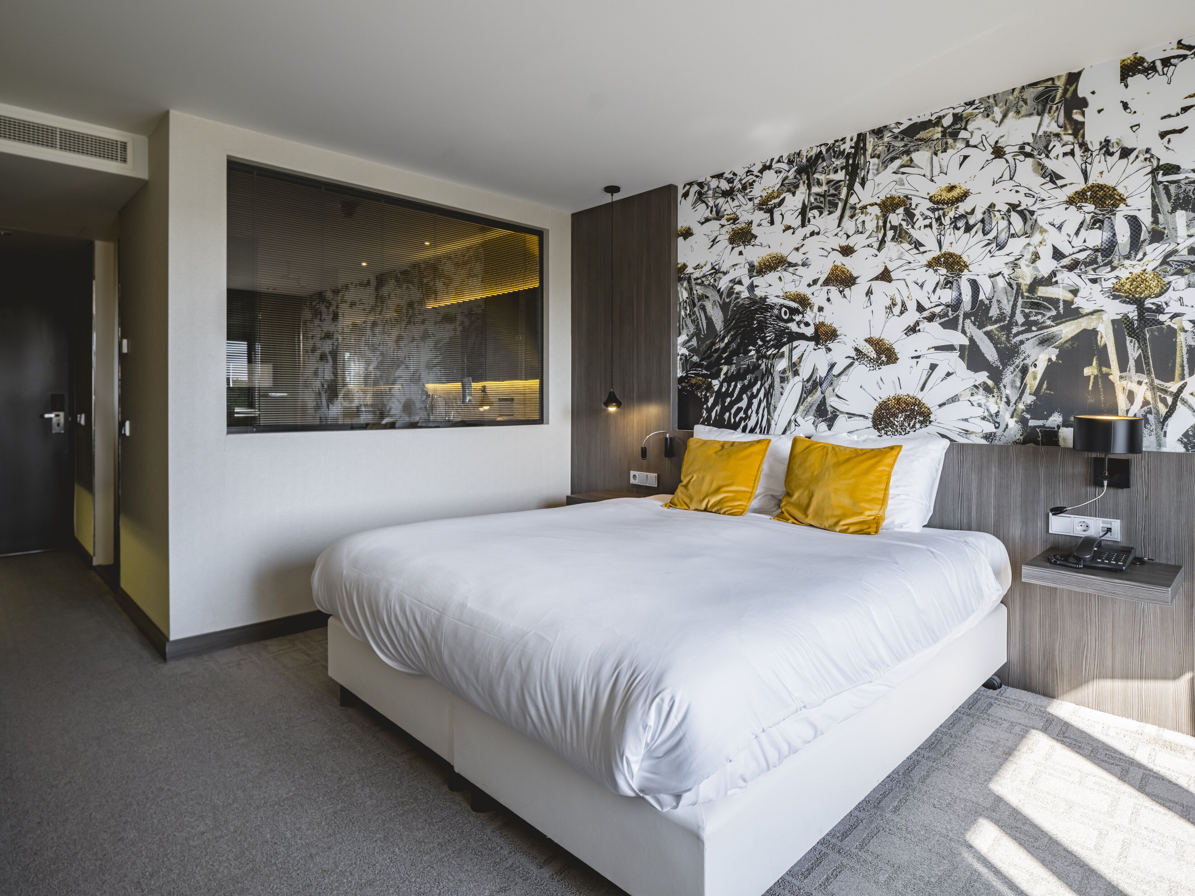 Quarto moderno com uma cama grande, janela com persianas, luminária na parede e papel de parede floral.