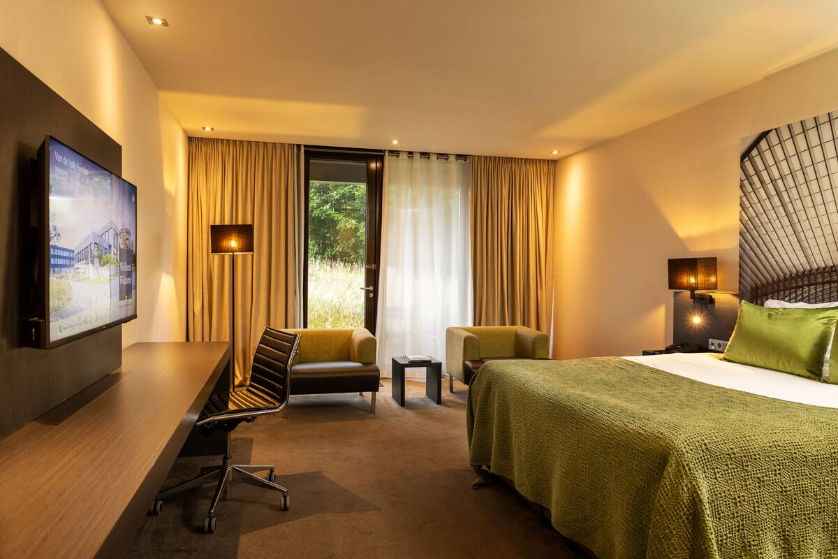Quarto de hotel com cama, duas poltronas, escrivaninha, televisão e uma janela grande com cortinas.