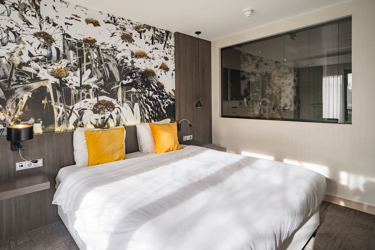Quarto moderno com cama grande, obra de arte na parede, mesa de cabeceira e janela com persianas.