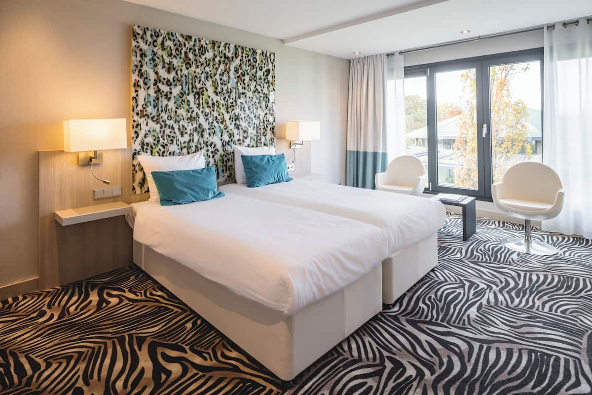 Quarto de hotel moderno com uma cama grande, duas mesas de cabeceira com luminárias, um tapete estampado com padrão de zebra e vista para o exterior por grandes janelas.
