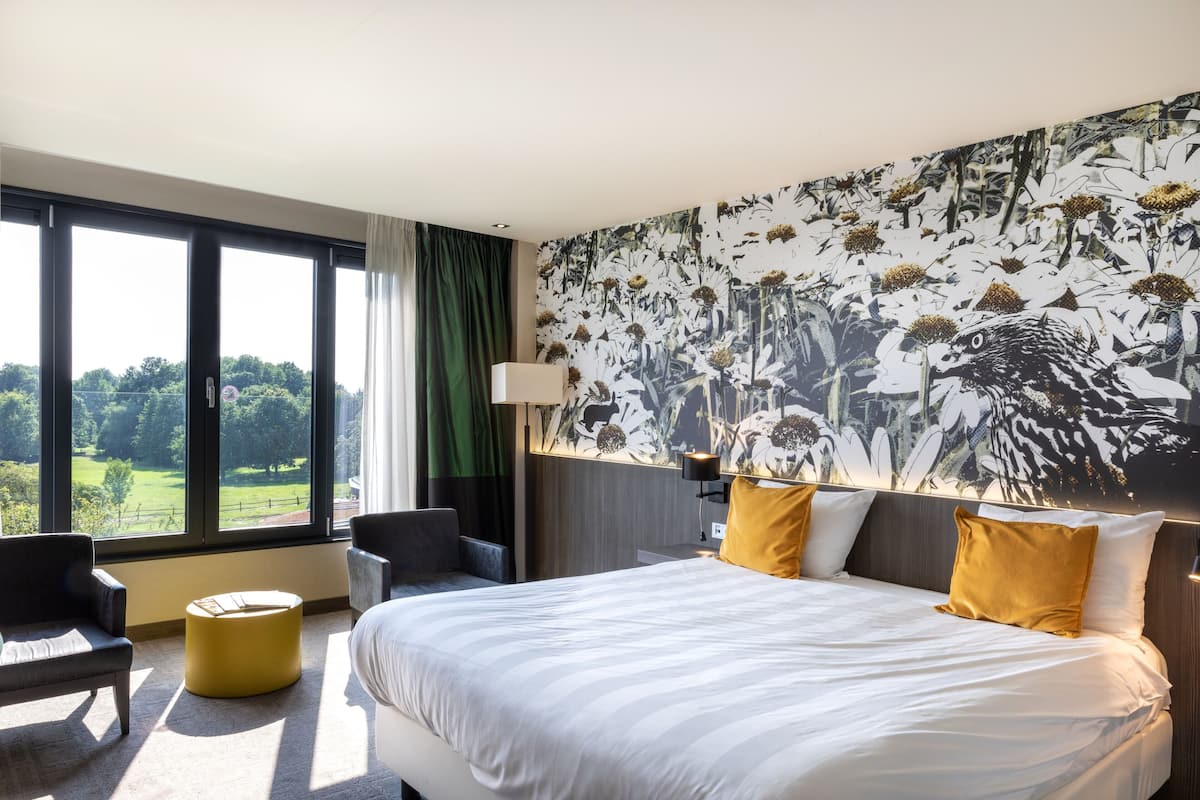 Quarto de hotel com cama grande, uma cadeira, um banquinho pequeno, mesa de cabeceira e uma janela ampla com vista para o verde.