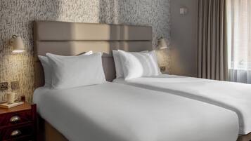 Quarto de hotel com duas camas, mesa de cabeceira com abajur, xícara e livro.