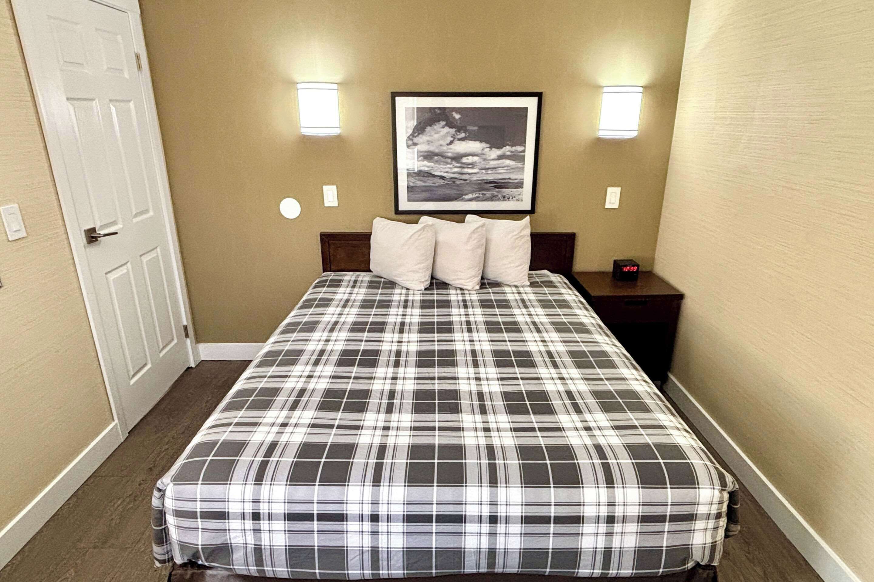 Quarto de hotel com uma cama, uma mesinha de cabeceira, um quadro pendurado na parede e duas luminárias fixadas na parede.