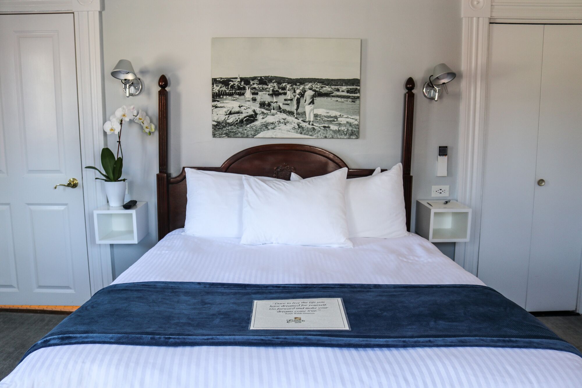 Cama bem arrumada com lençóis brancos e um toque azul, cabeceira de madeira, quadro pendurado na parede, abajures e uma pequena mesa de cabeceira com uma planta.