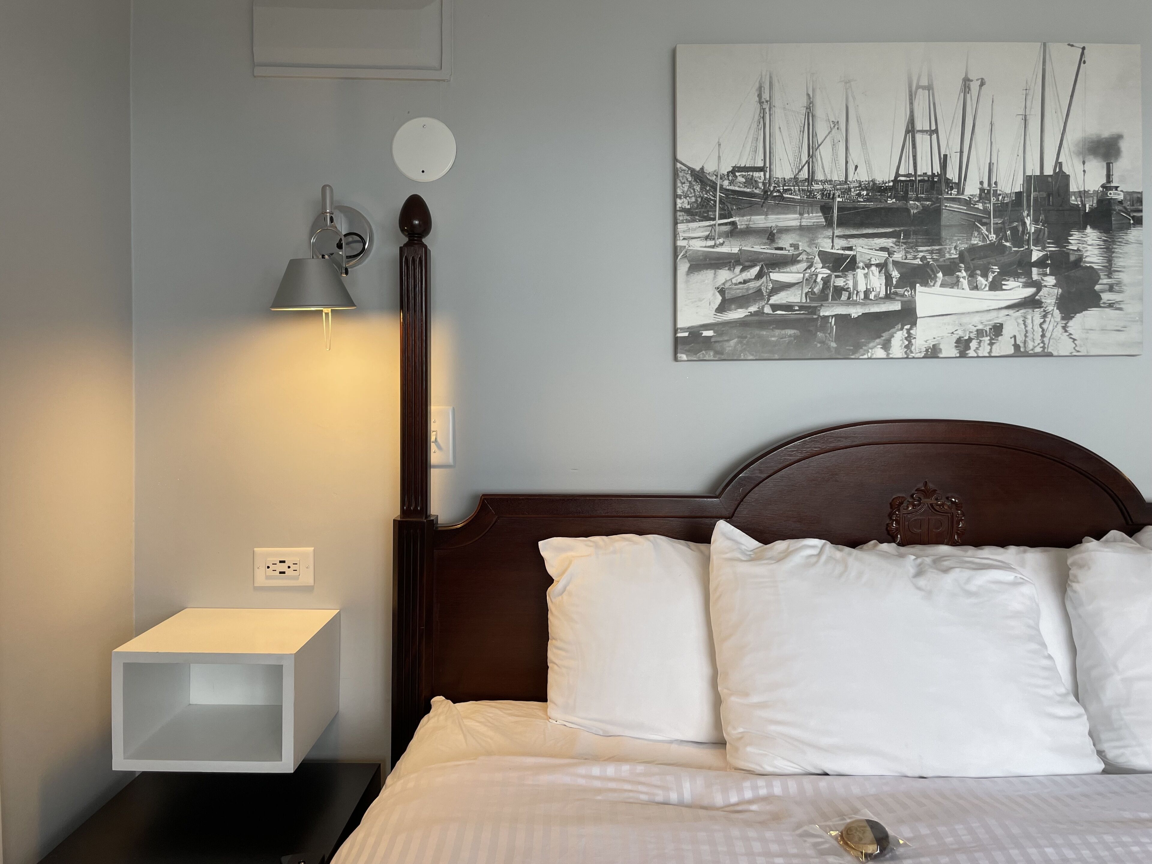Cama bem arrumada com lençóis brancos, cabeceira de madeira, mesa de cabeceira com abajur e um quadro na parede retratando uma paisagem portuária.