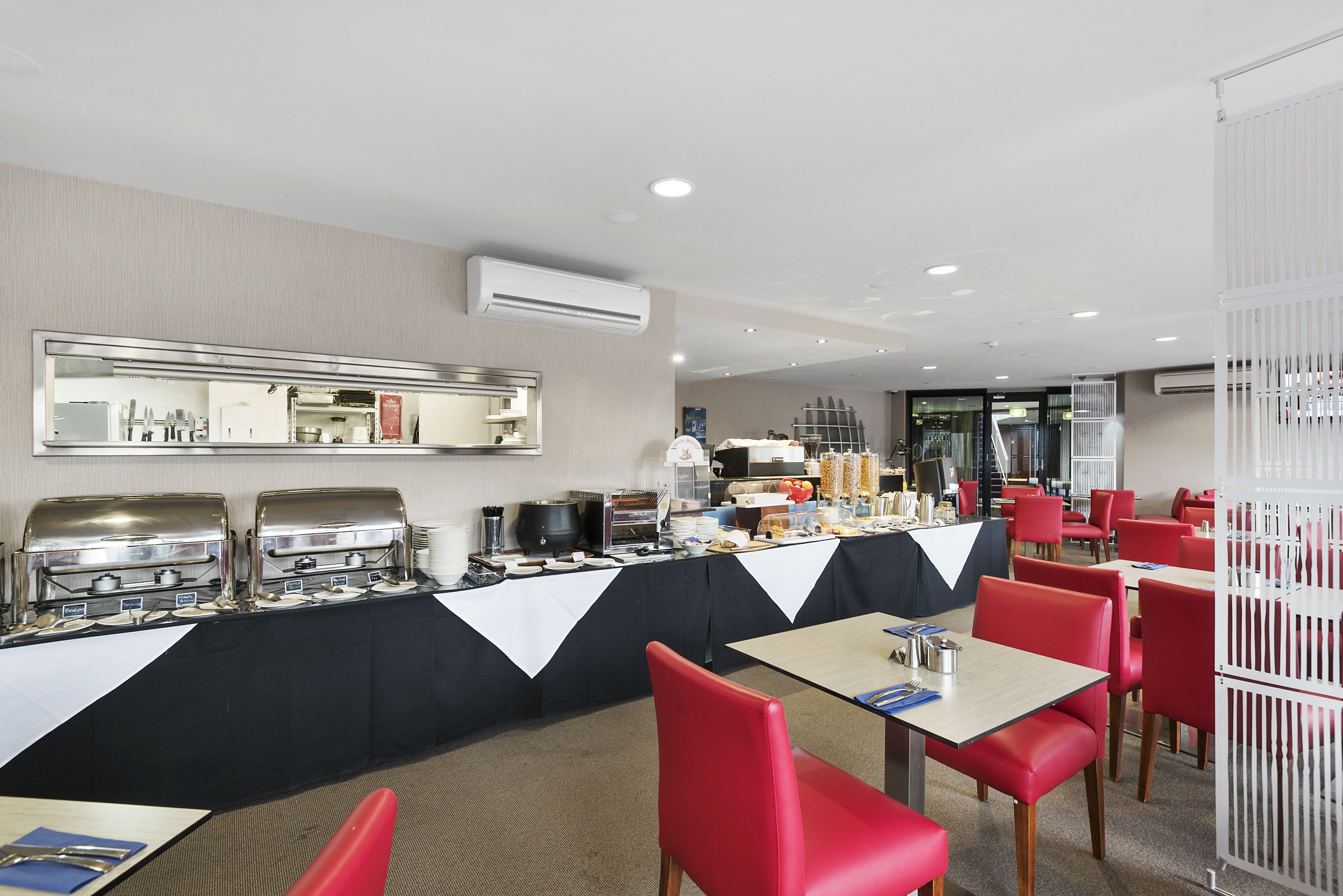 Disposição de buffet com várias estações de comida, mesas e cadeiras vermelhas numa área de refeições.