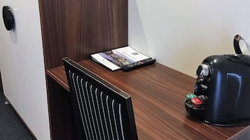 Quarto de hotel com uma escrivaninha de madeira, uma cadeira preta, uma televisão sobre um suporte e uma mala preta na escrivaninha.