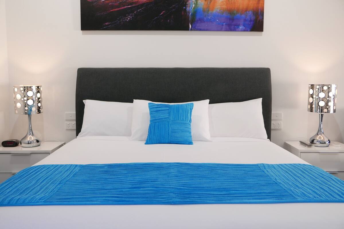 Uma cama bem arrumada com travesseiro e colcha azuis, com duas mesas de cabeceira com luminárias, em um quarto de hotel moderno.