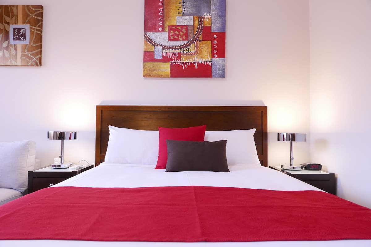 Uma cama bem arrumada com um cobertor vermelho, duas mesas de cabeceira com luminárias e um quadro abstrato colorido na parede.
