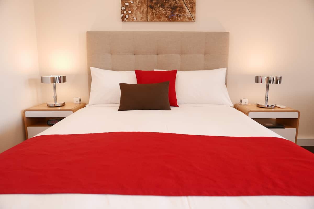 Uma cama bem arrumada com colcha vermelha e branca, duas mesas de cabeceira com luminárias e uma cabeceira com design texturizado.