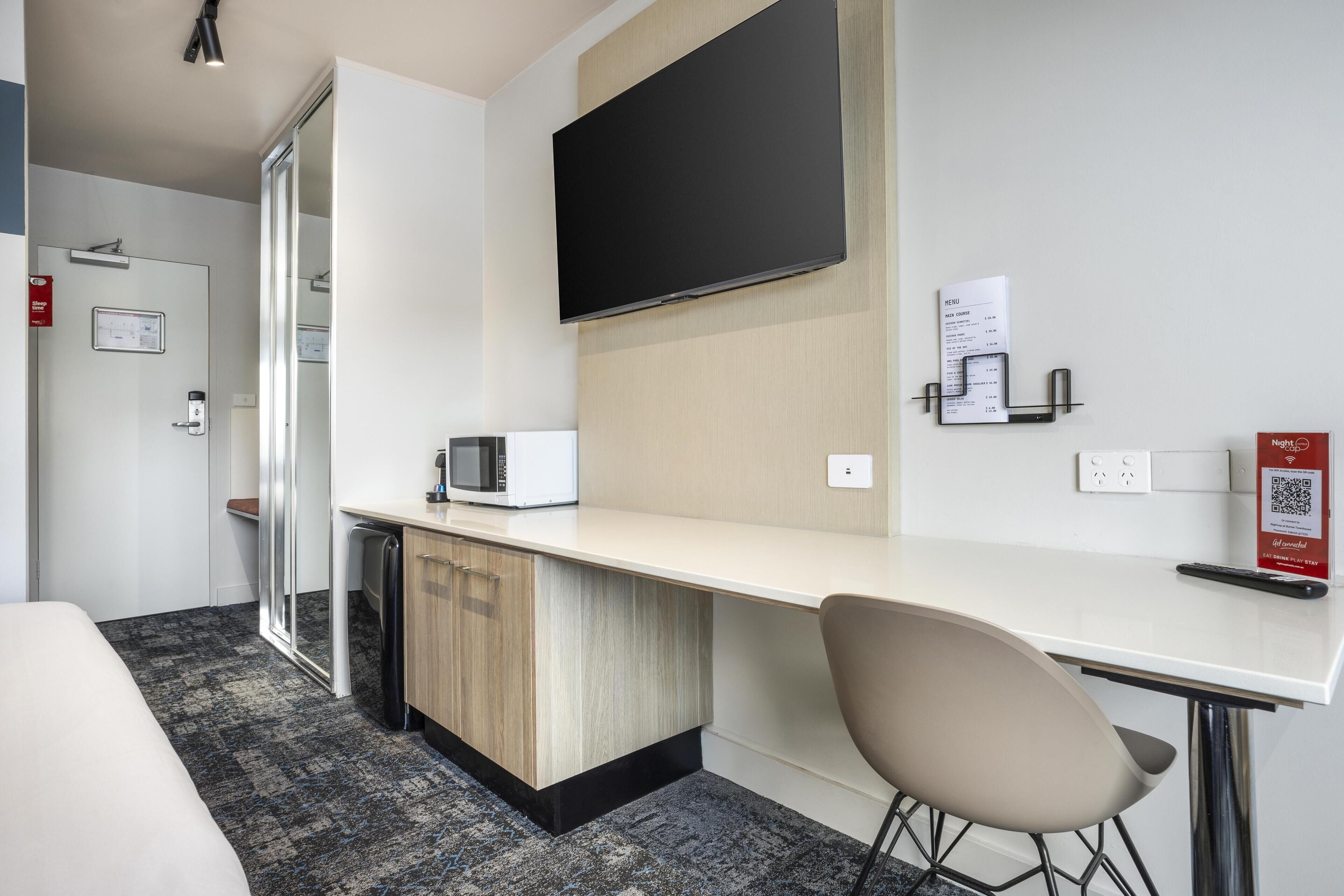 Quarto de hotel com cama, escrivaninha com cadeira, micro-ondas, televisão de tela plana, porta com painel de vidro e quadro de cardápio fixado na parede.