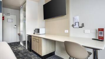 Quarto de hotel com cama, escrivaninha com cadeira, micro-ondas, televisão de tela plana, porta com painel de vidro e quadro de cardápio fixado na parede.