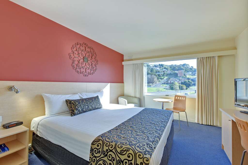 Quarto de hotel com uma cama grande, uma escrivaninha, uma cadeira e uma televisão.