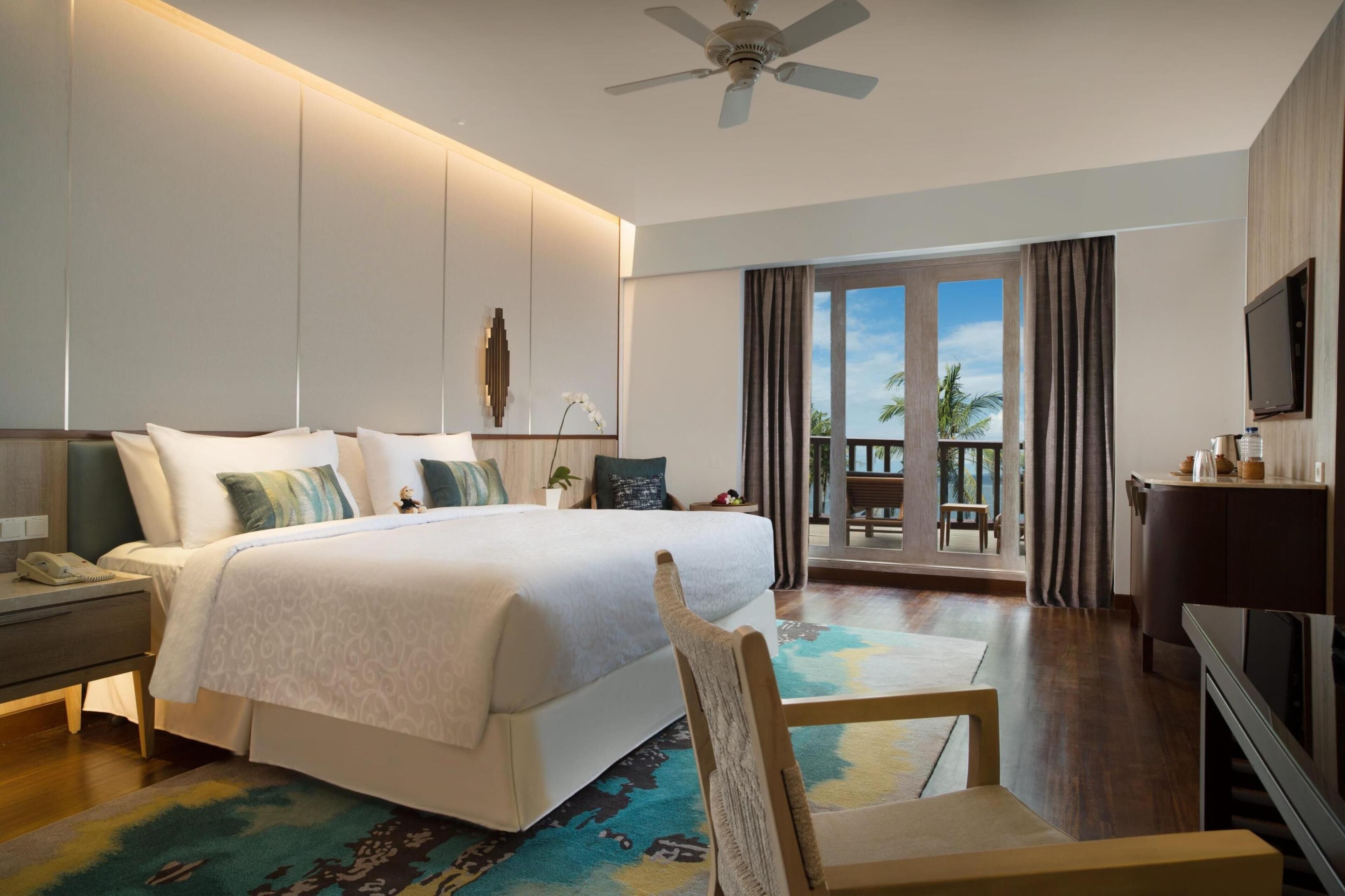 Quarto de hotel com uma cama grande, uma escrivaninha com telefone, televisão e varanda com vista para palmeiras.