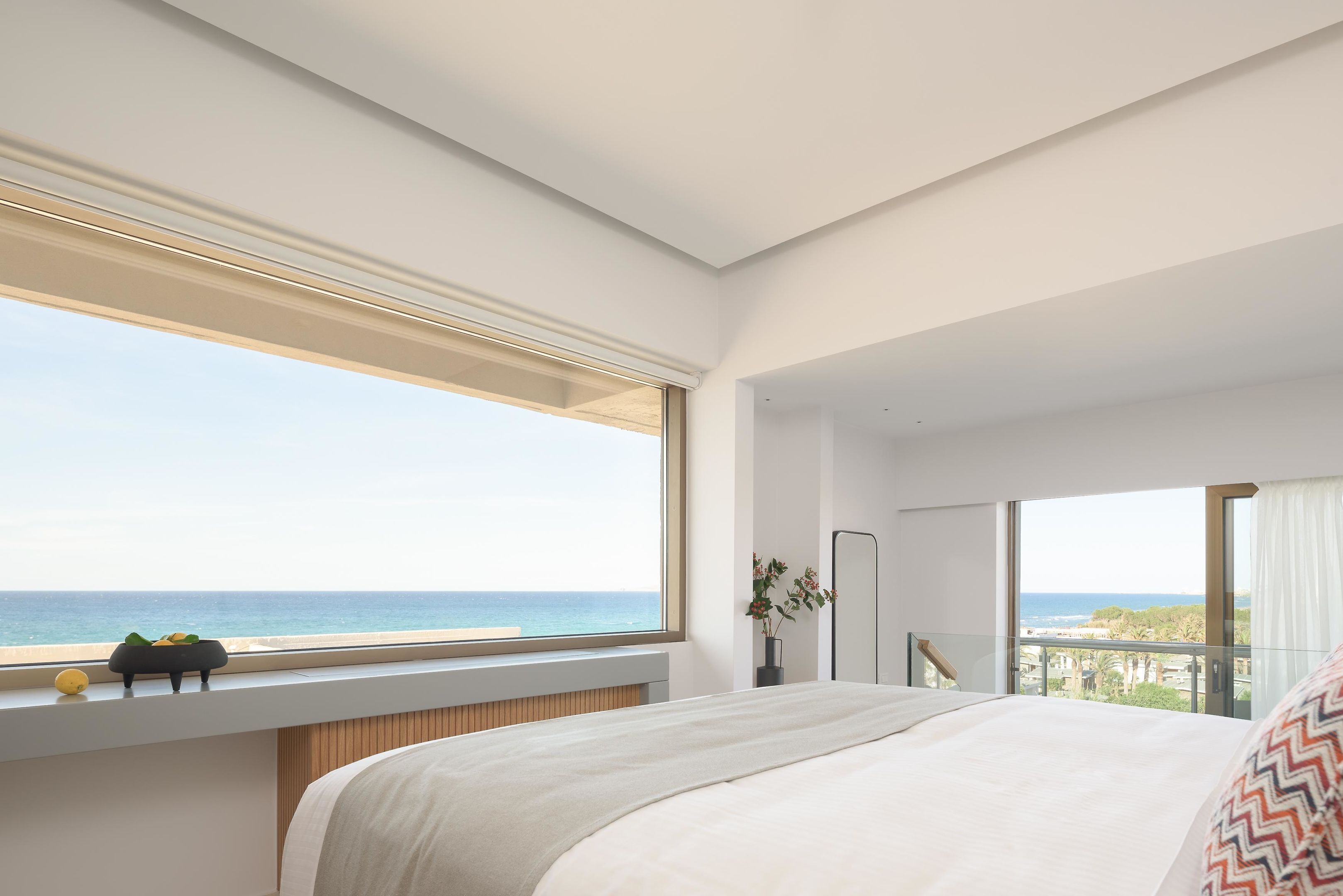 Quarto com uma grande janela com vista para o mar, uma cama e uma pequena mesa com uma planta.