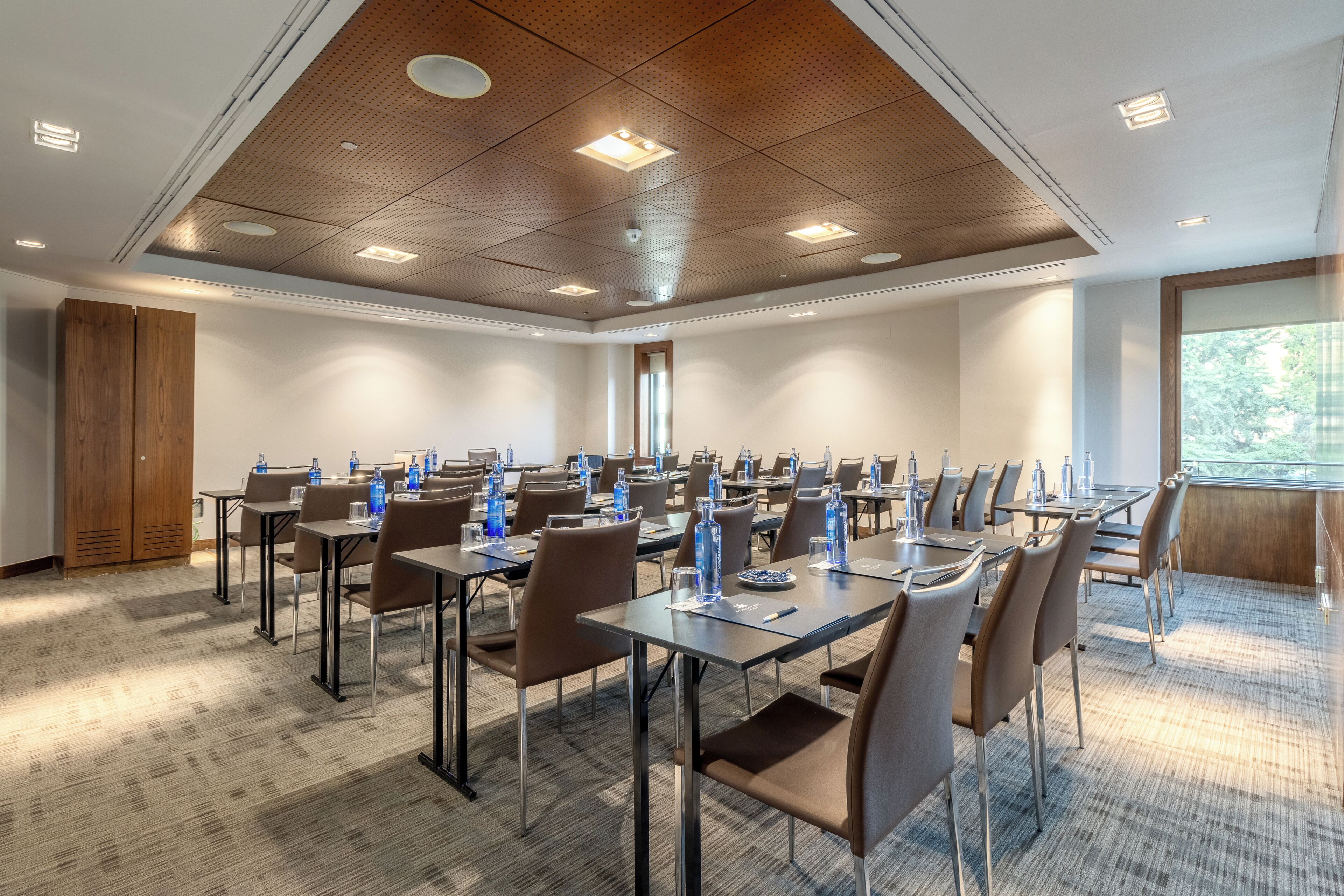 Uma sala de conferências com várias mesas e cadeiras, equipada com garrafas de água e copos, e um grande janelão que ilumina o espaço com luz natural.