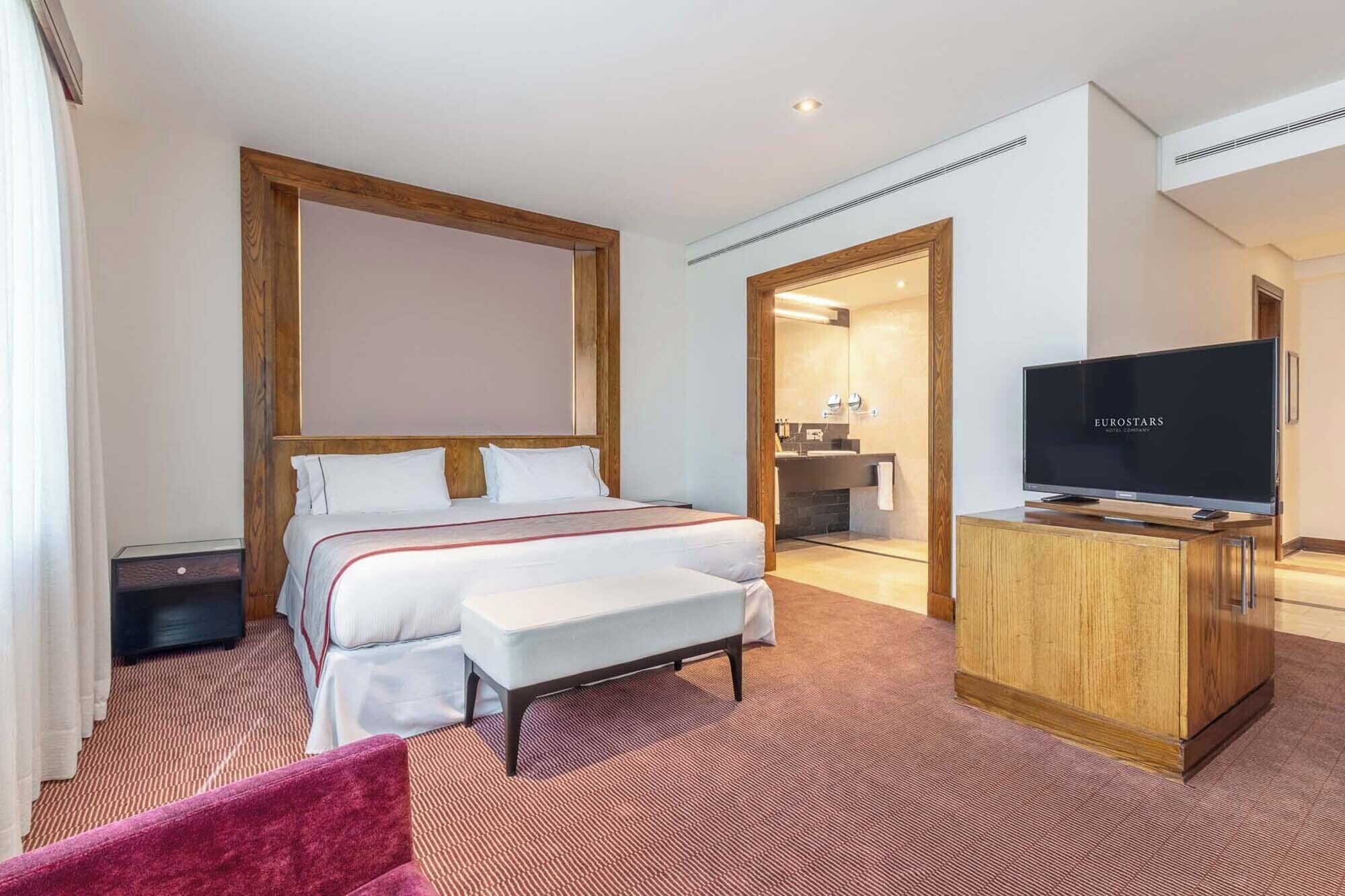 Quarto de hotel com uma cama grande, televisão, armário de madeira e banheiro visível por uma porta aberta.