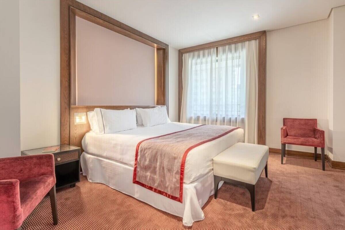 Quarto de hotel com uma cama grande, mesa de cabeceira, uma cadeira e uma janela com cortinas.