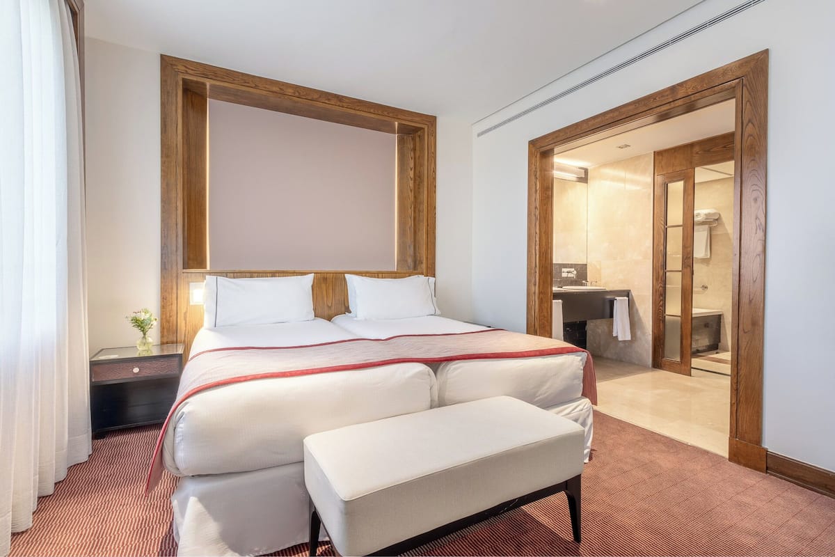 Quarto de hotel com uma cama grande, um banco, uma mesinha de cabeceira e um banheiro visível através de uma porta aberta.