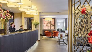 Uma área de recepção de hotel com um balcão escuro, várias luminárias e uma área de estar com cadeiras laranja.