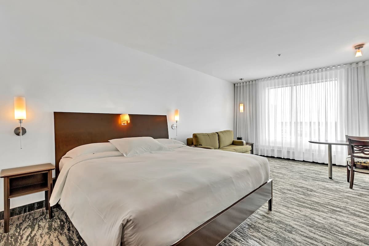 Quarto de hotel com uma cama grande, uma mesinha de cabeceira, um pequeno sofá, uma mesa de jantar e cortinas.