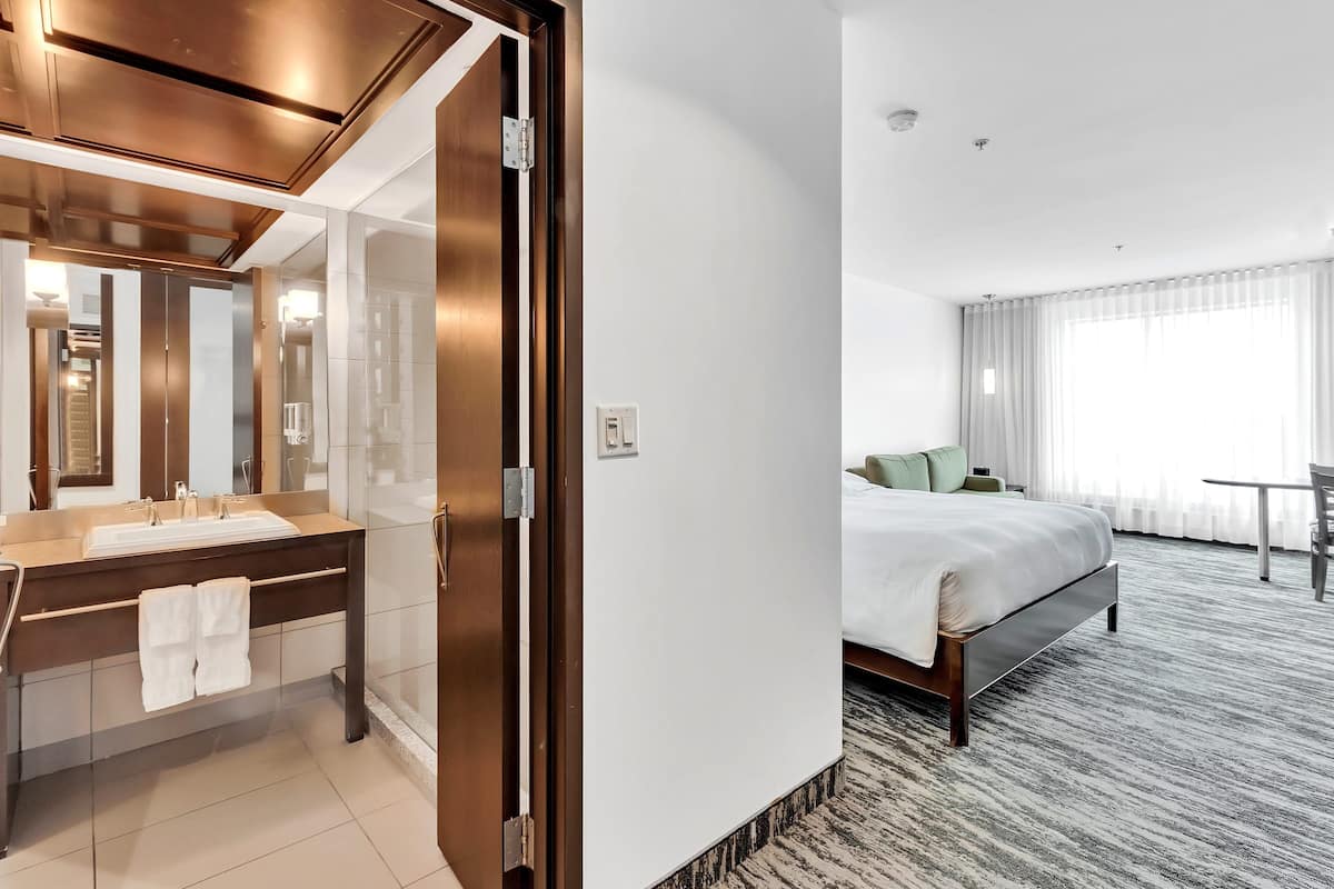 Quarto de hotel moderno com banheiro que inclui um espelho grande, pia e porta-toalhas. O quarto possui uma cama, uma mesa pequena e uma janela com cortinas.